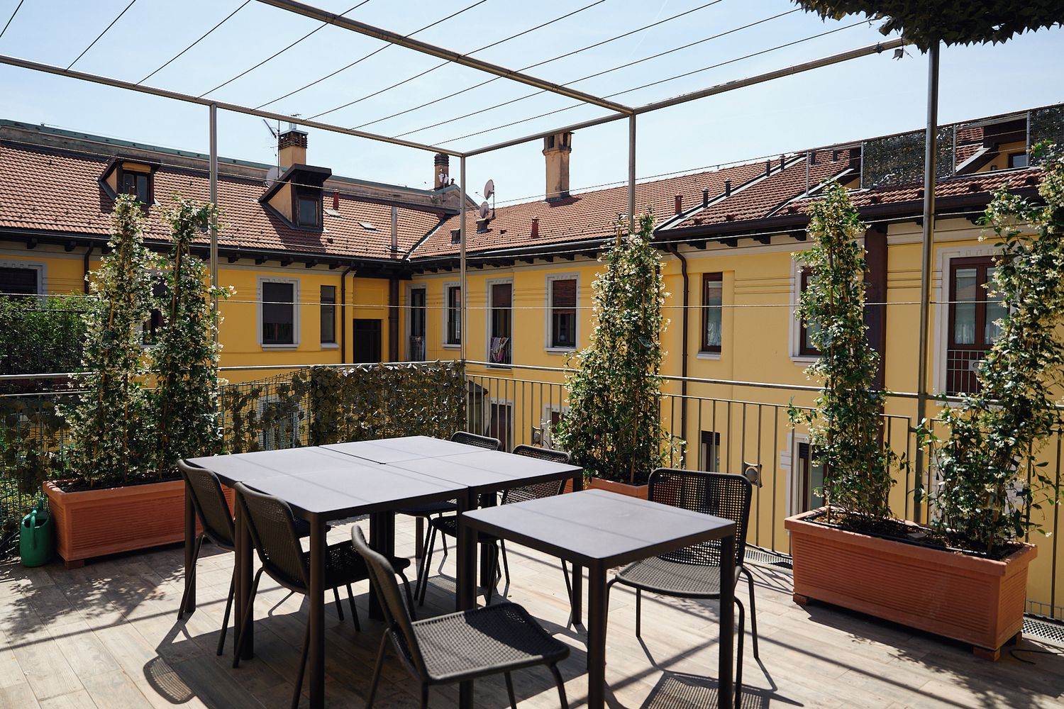 Um pátio externo com mesa e cadeiras, rodeado por plantas em vasos e um edifício amarelo com telhado de azulejos.
