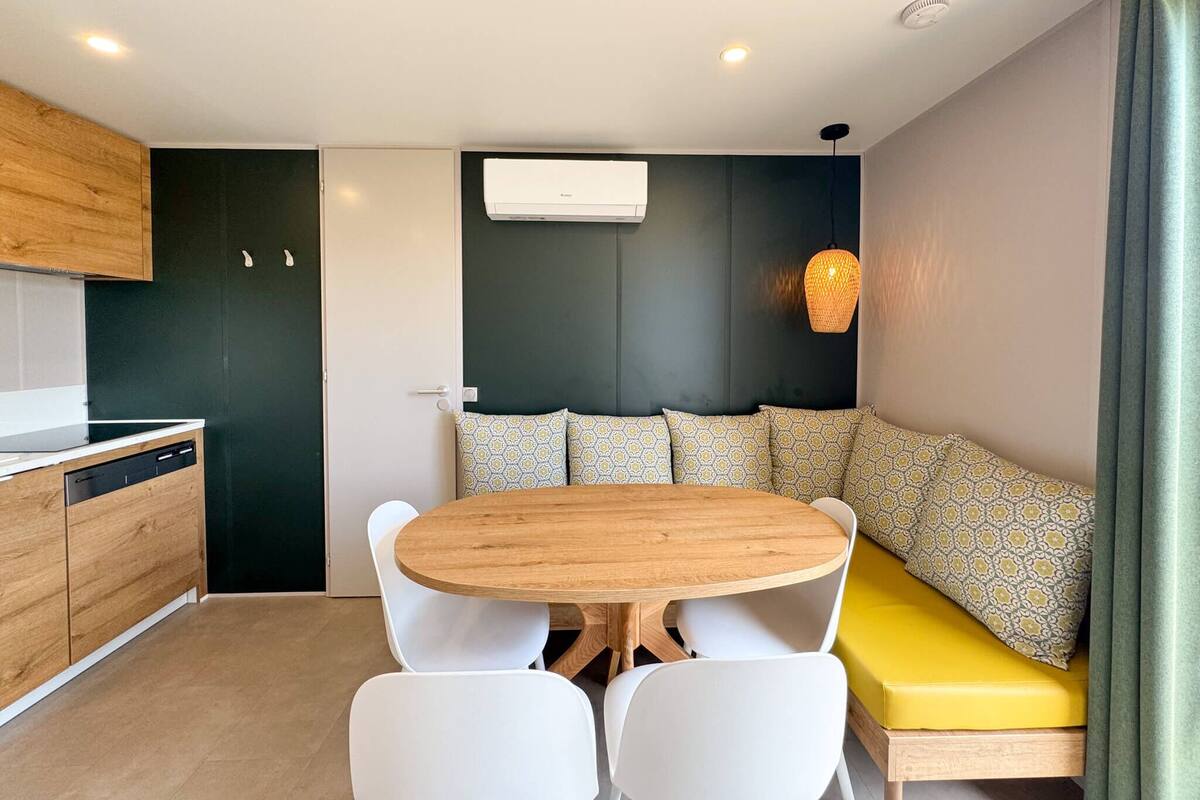 Um espaço compacto com cozinha americana, mesa de jantar com cadeiras, sofá com almofadas estampadas e luminária pendente.