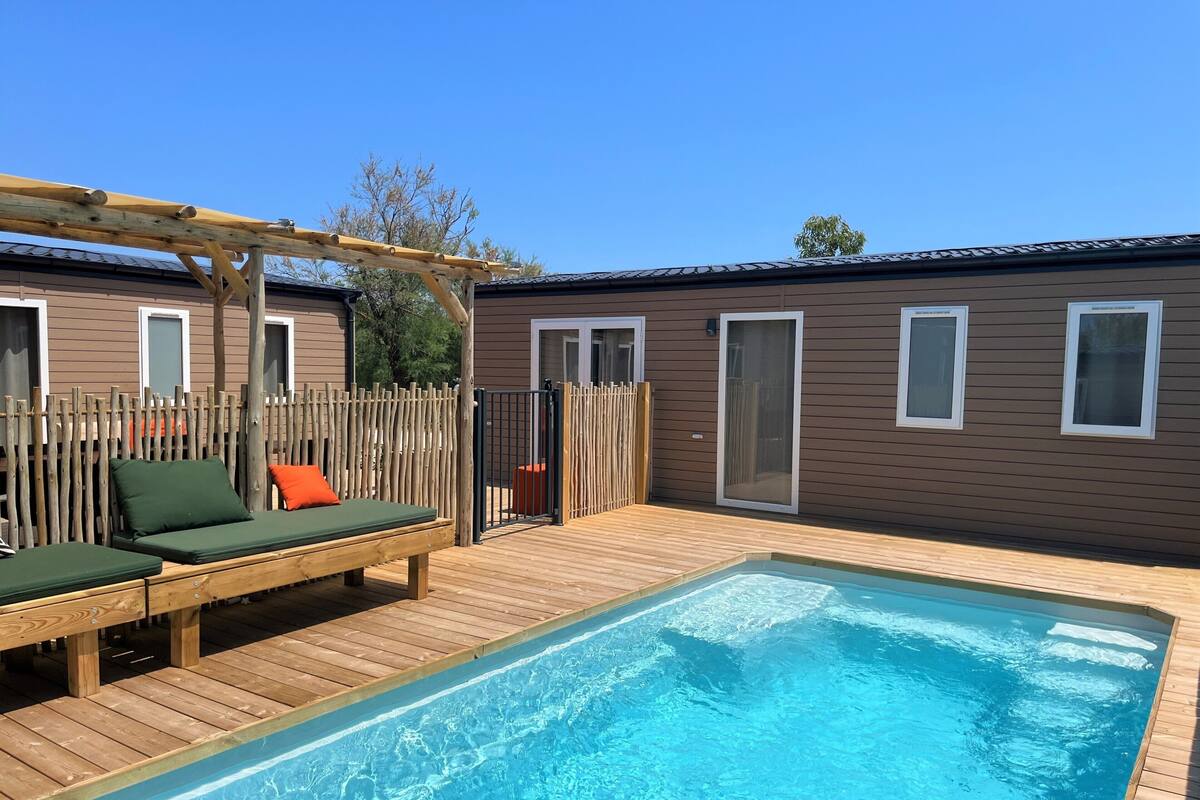 Um deck de madeira com piscina, uma pérgula de madeira e uma área cercada com um banco e almofadas.