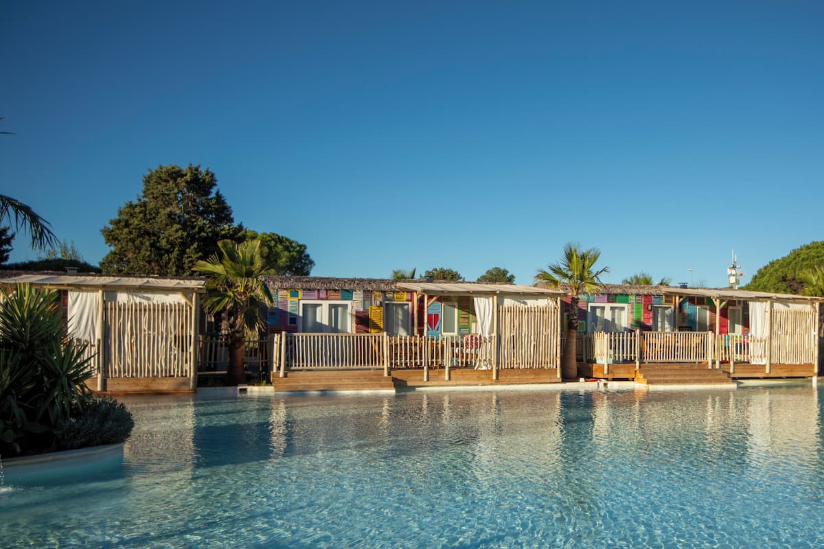 Uma área de piscina com várias cabanas pequenas e coloridas, cada uma com um deque de madeira e um espaço de estar coberto.