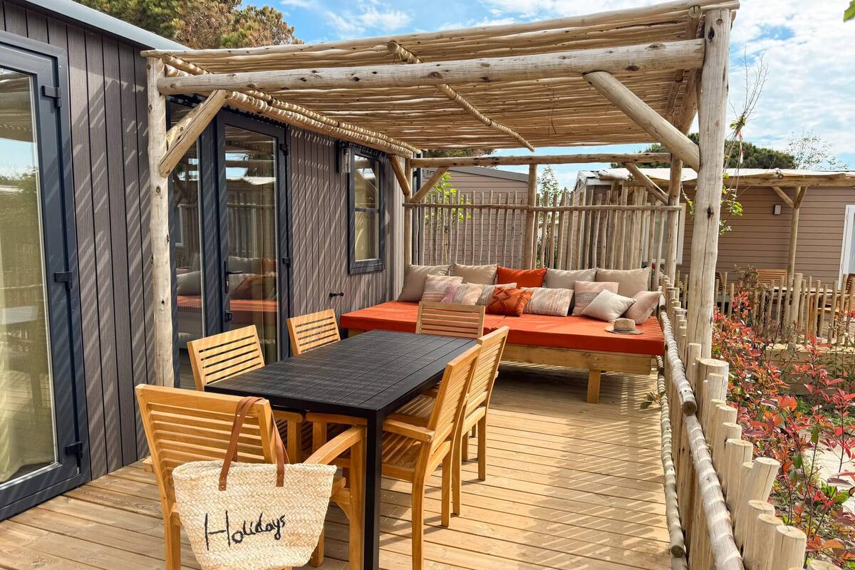 Um deck de madeira com mesa e cadeiras, um sofá com almofadas e uma bolsa trançada com a etiqueta 'Férias'.