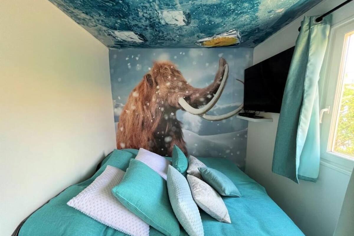 Quarto com um mural de um mamute, cama com roupa de cama verde-água e janela com cortinas.