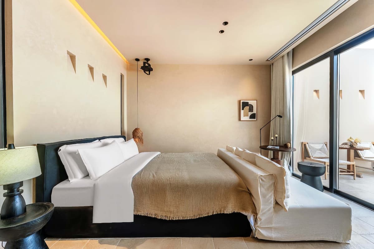 Quarto de hotel moderno com uma cama grande, uma mesinha de cabeceira, um abajur, vista para o exterior e um quadro na parede.