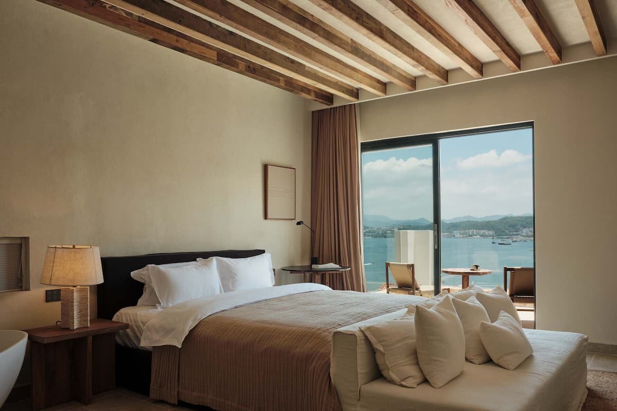 Quarto de hotel com uma cama, mesa de cabeceira com abajur, vista para o mar e varanda com mesa e cadeiras.