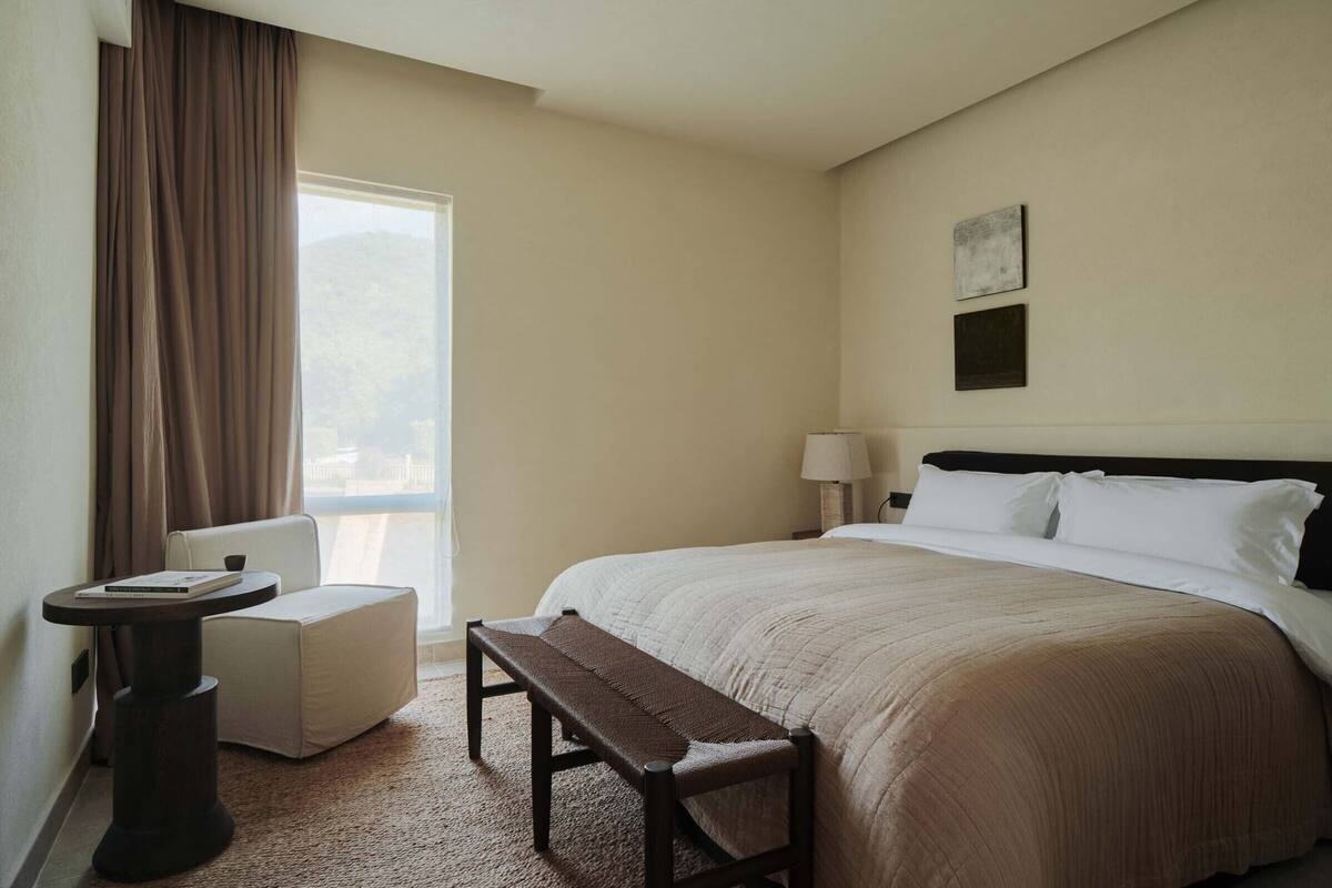 Quarto de hotel com cama, um banco, uma mesinha, uma cadeira, um abajur e uma janela com cortinas.