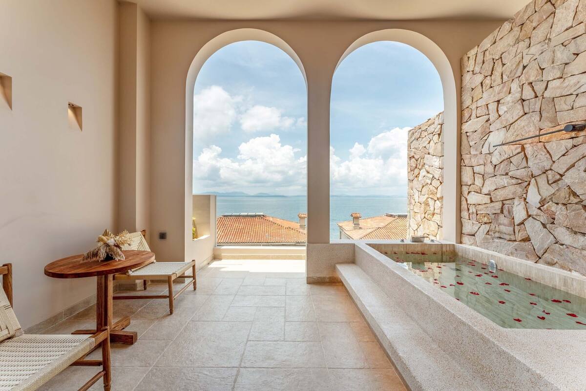Uma varanda com parede de pedra, uma piscina com pétalas vermelhas e vista para o mar.