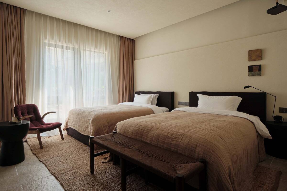 Quarto de hotel com duas camas, uma cadeira, um banco, uma escrivaninha e uma janela com cortinas.