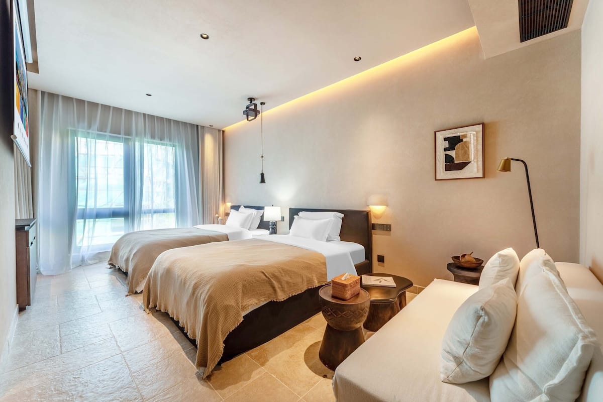 Quarto de hotel com duas camas, um sofá, uma mesa de cabeceira, uma luminária e uma janela com cortinas.