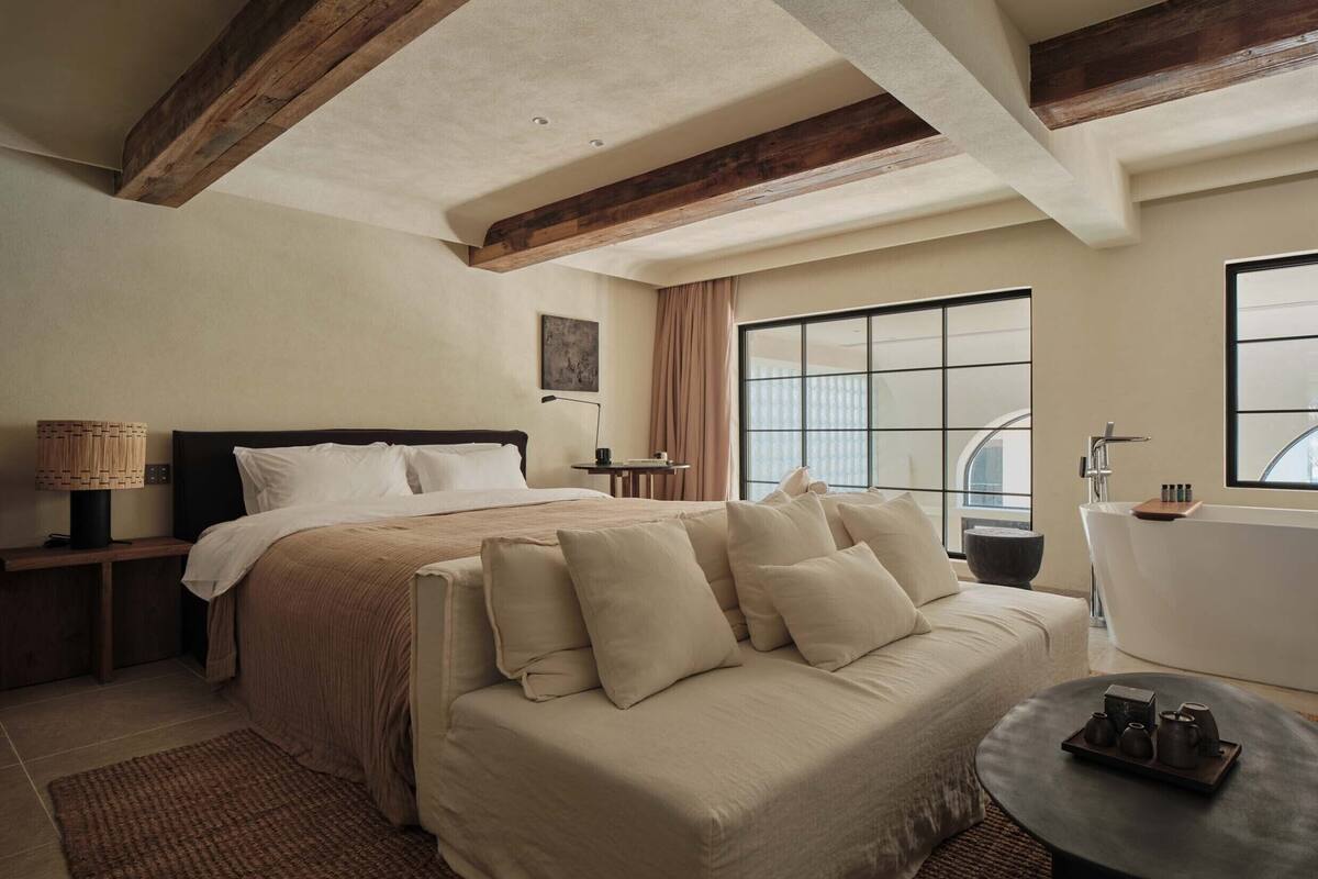 Quarto de hotel com uma cama grande, um sofá, uma mesa de centro redonda, uma escrivaninha com abajur, uma janela ampla e vista para a cidade.