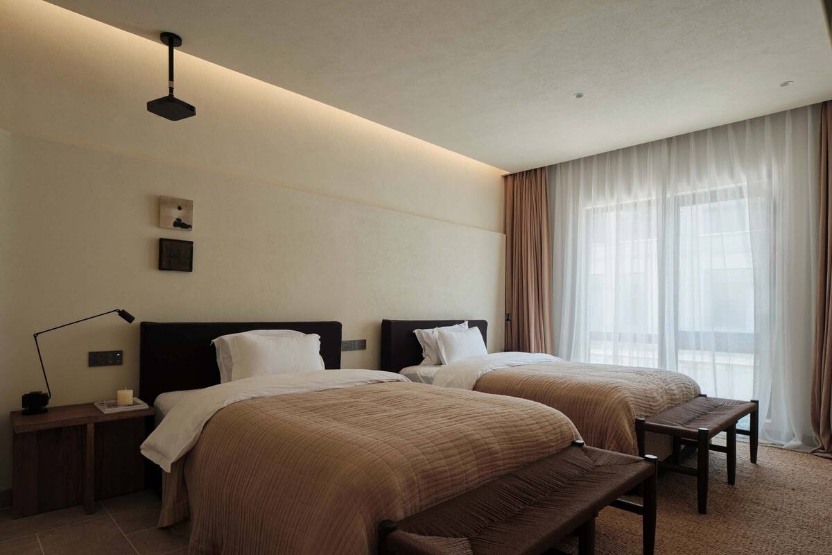 Quarto de hotel com duas camas, uma mesinha de cabeceira com luminária, uma pequena mesa e um banco.