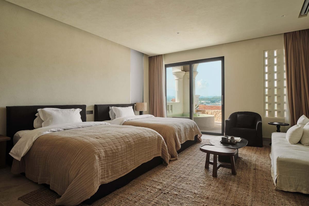 Quarto de hotel com duas camas, uma mesinha, uma cadeira e varanda com vista.