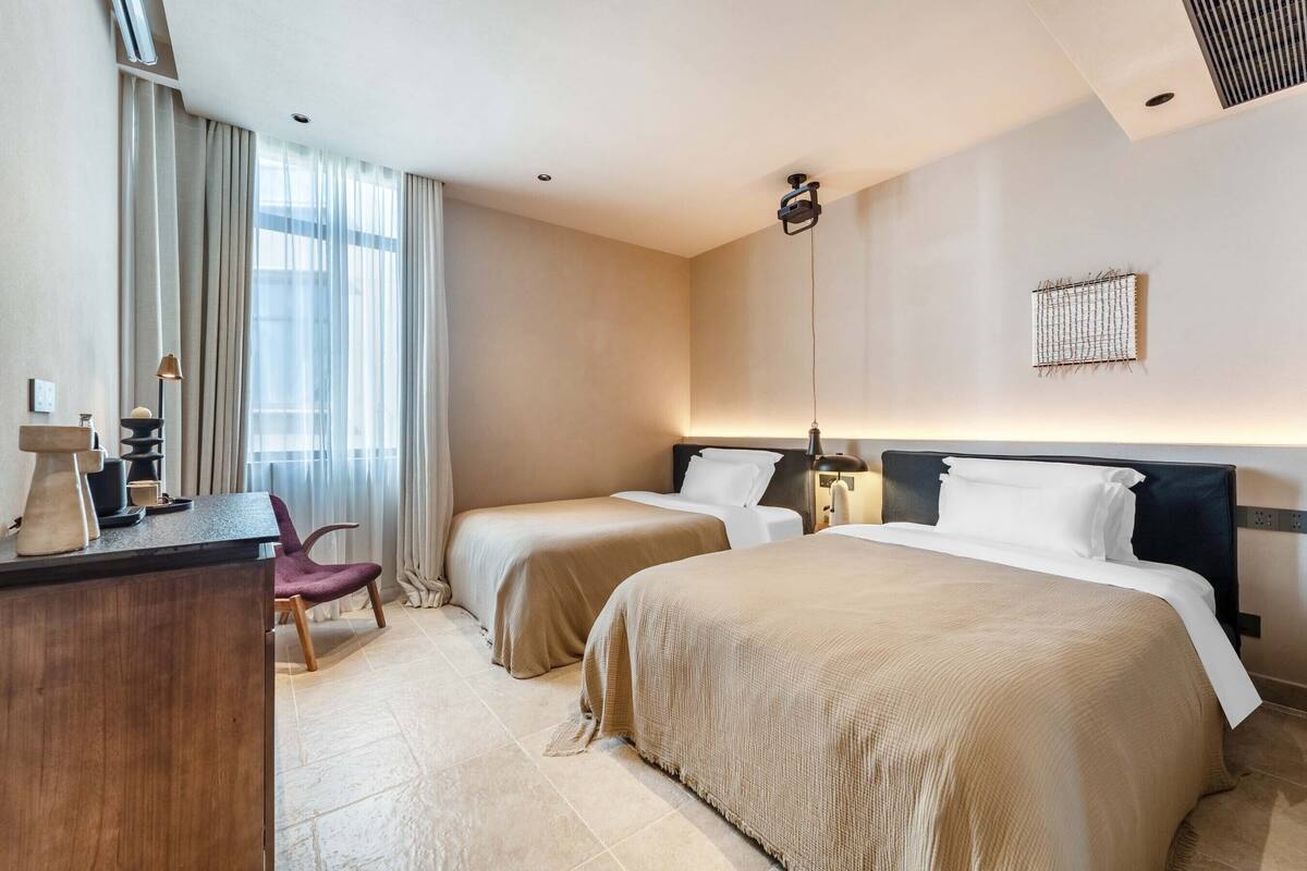 Quarto de hotel com duas camas, uma escrivaninha, uma cadeira, um abajur e uma janela grande com cortinas.