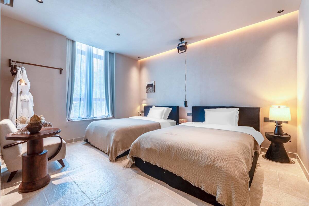 Quarto de hotel com duas camas, área de estar com mesa e janela com cortinas.