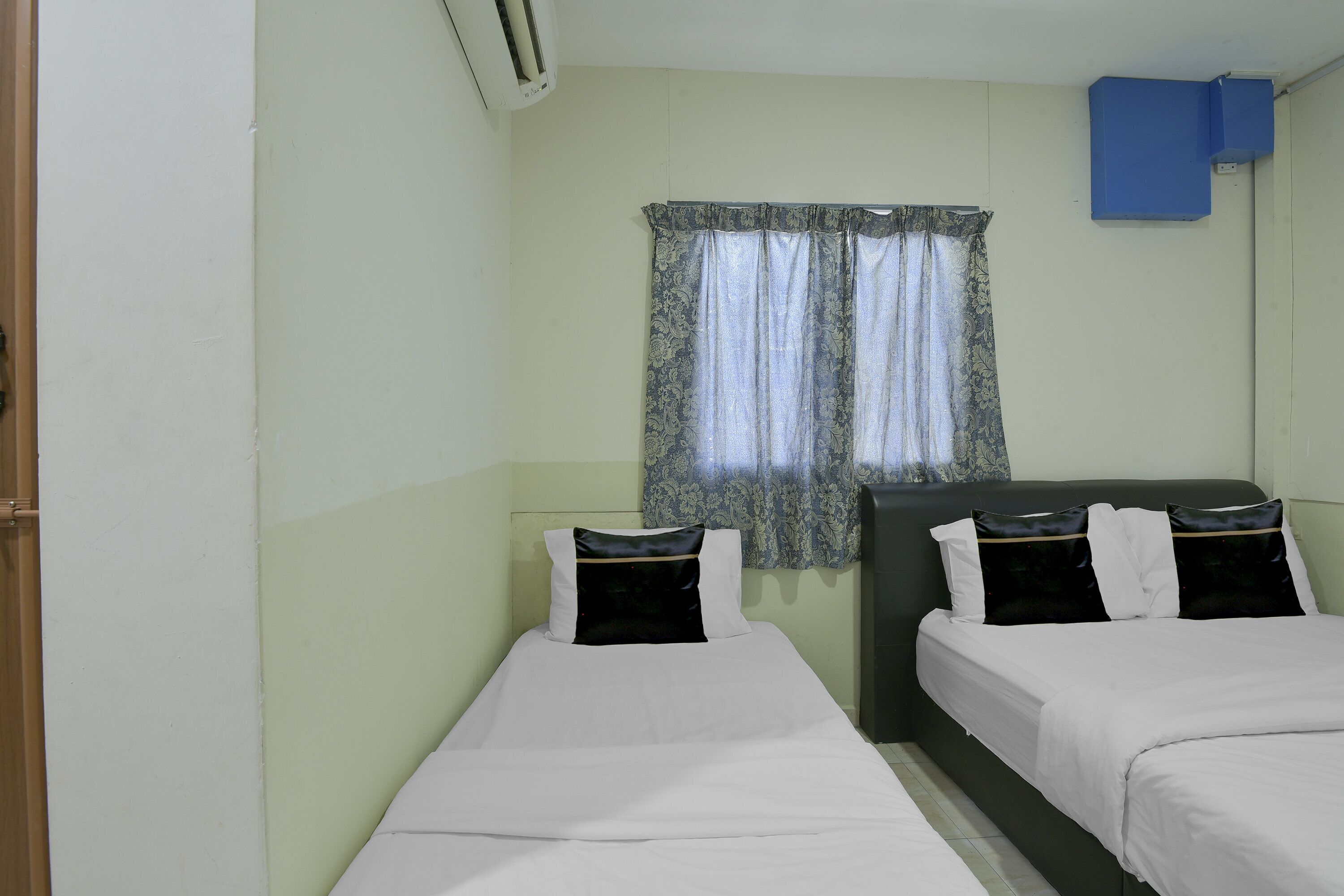 Quarto com duas camas, uma janela com cortinas, ar condicionado fixado na parede e um objeto azul na parede.
