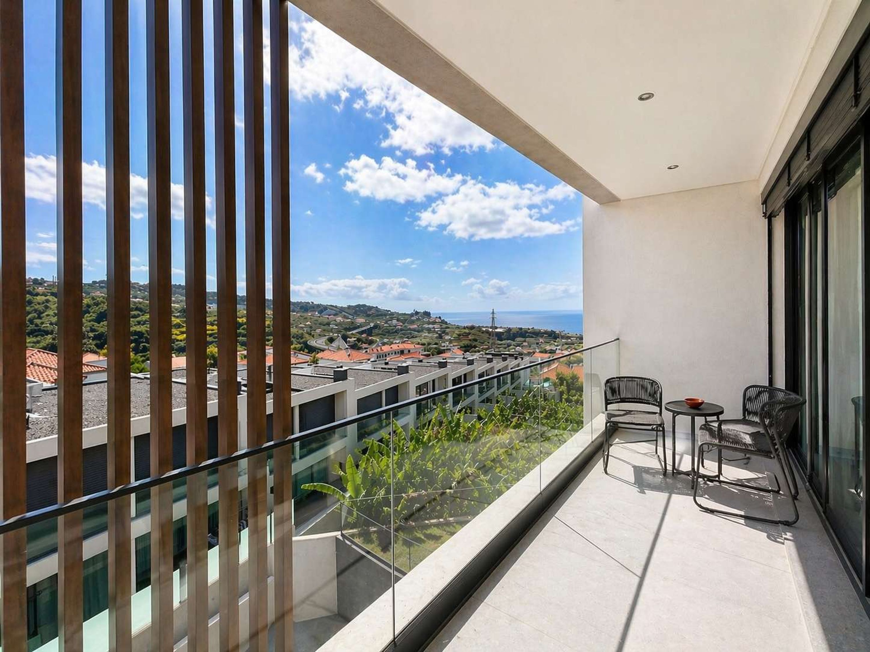 Uma varanda com guarda-corpo de vidro, uma mesa com cadeiras e vista para a paisagem costeira.