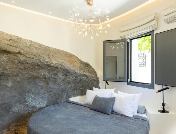 Quarto com uma grande parede de pedra, cama com roupa de cama cinzenta, lustre moderno, janela com cortinas e mesa de cabeceira com abajur.
