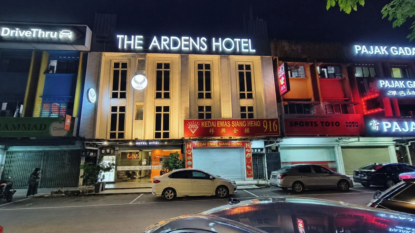 O Hotel Ardens com uma placa visível, ao lado de um DriveThru e um restaurante Kedi Emas Siang Heng.