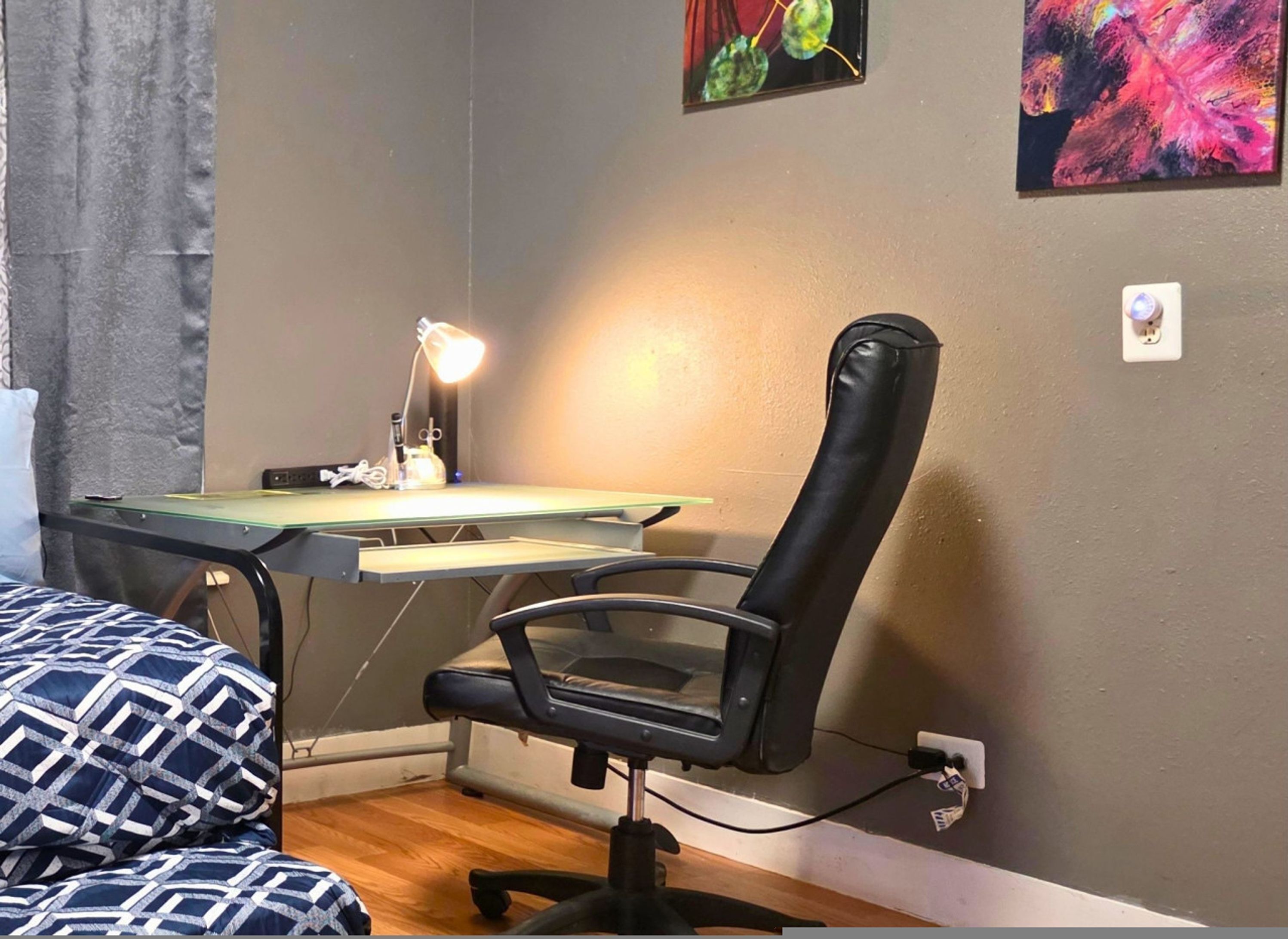 Um quarto com uma cama, uma escrivaninha com luminária, uma cadeira e uma parede com duas pinturas.