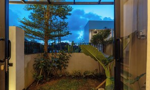 Visão através de uma porta de vidro de um edifício, com uma árvore, cerca e outro edifício ao fundo.