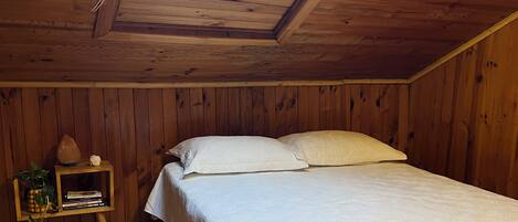 Quarto com claraboia, paredes de madeira, cama com roupa de cama branca e uma pequena mesa de cabeceira com livros e uma planta.