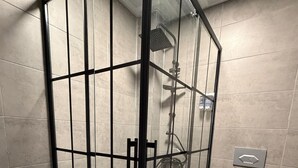 Um chuveiro com box de vidro e moldura preta, um vaso sanitário e um painel de controle fixado na parede.