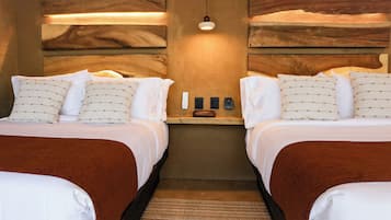 Quarto de hotel com duas camas, uma mesinha e luminária fixada na parede.