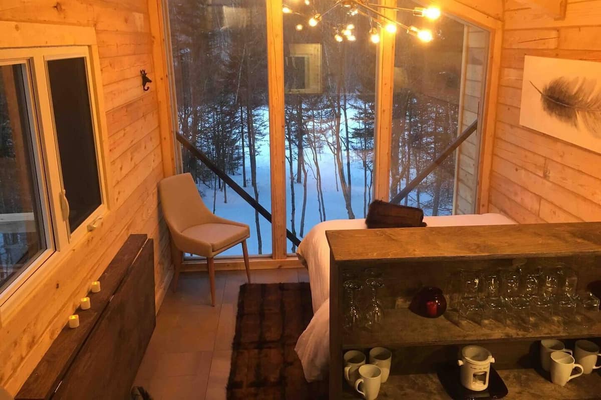 Um quarto de cabana aconchegante, com uma cama, uma poltrona, uma janela com vista para uma floresta coberta de neve e uma estante com copos e canecas.