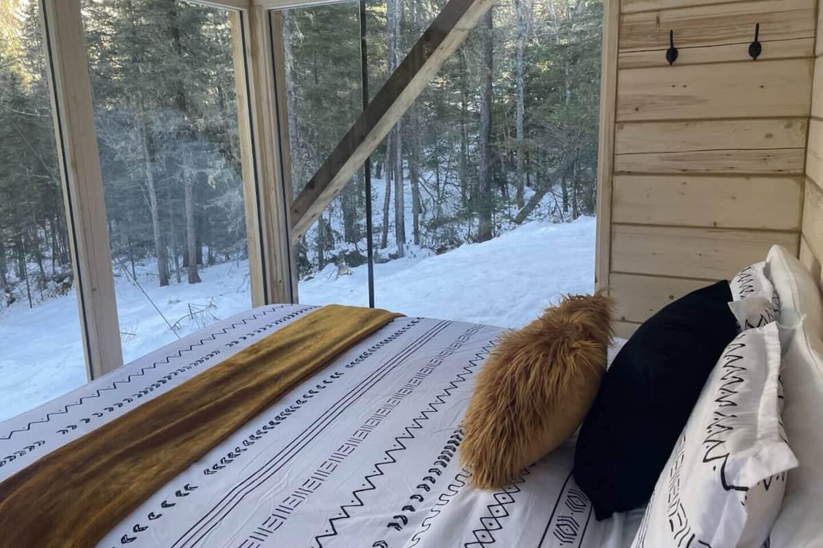 Uma cabana com uma cama de frente para uma vista nevada da floresta através de grandes janelas.
