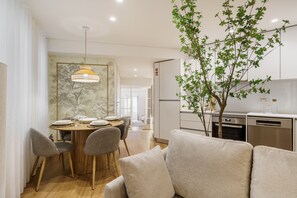 Cozinha moderna com eletrodomésticos de aço inoxidável, área de jantar com mesa e cadeiras redondas, e sala de estar com sofá e planta.