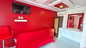 Um sofá vermelho, uma televisão na parede, um espelho e uma cadeira vermelha num quarto com paredes brancas e chão em azulejos.