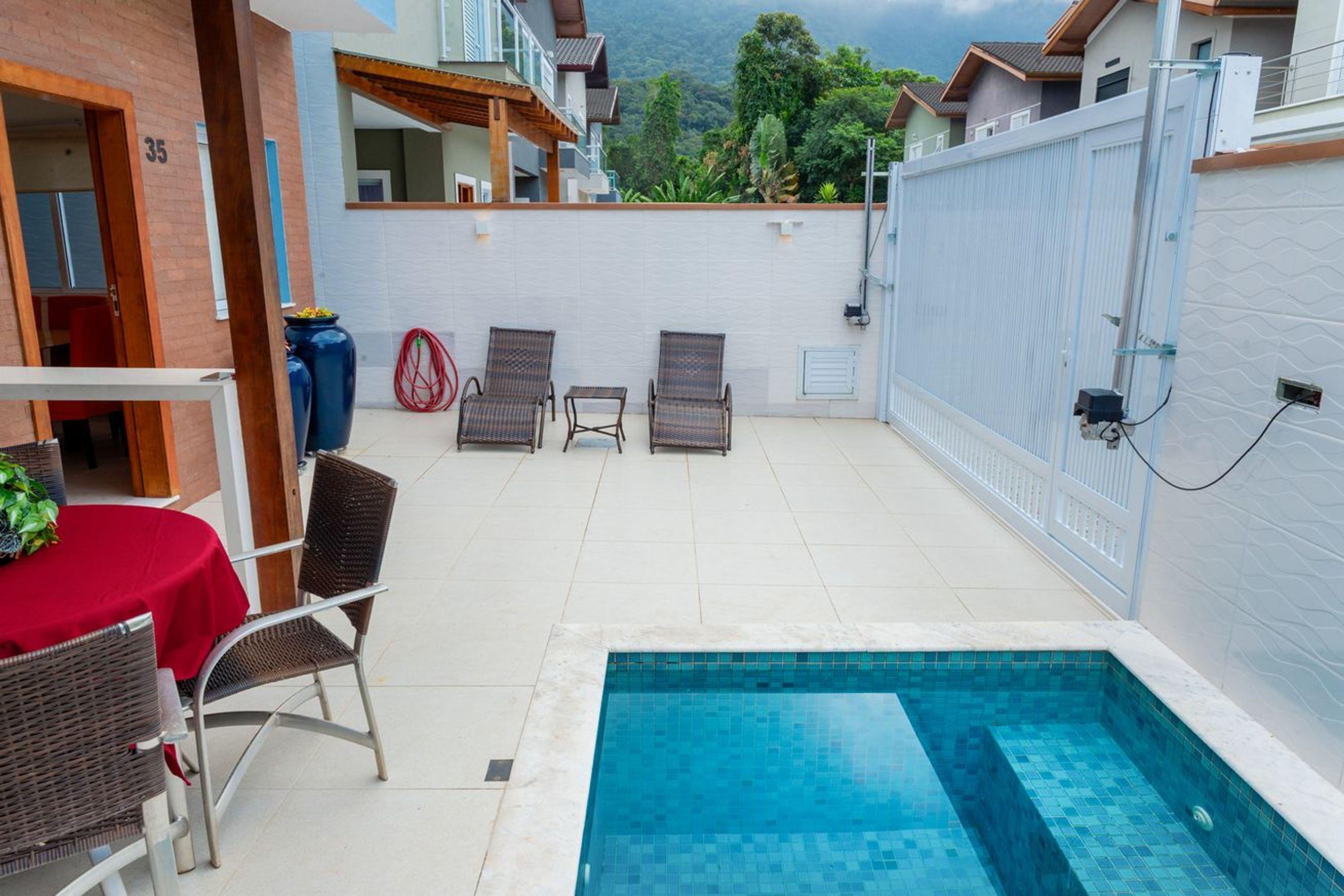 Piscina com deck revestido, cadeiras de vime e uma mesa vermelha.