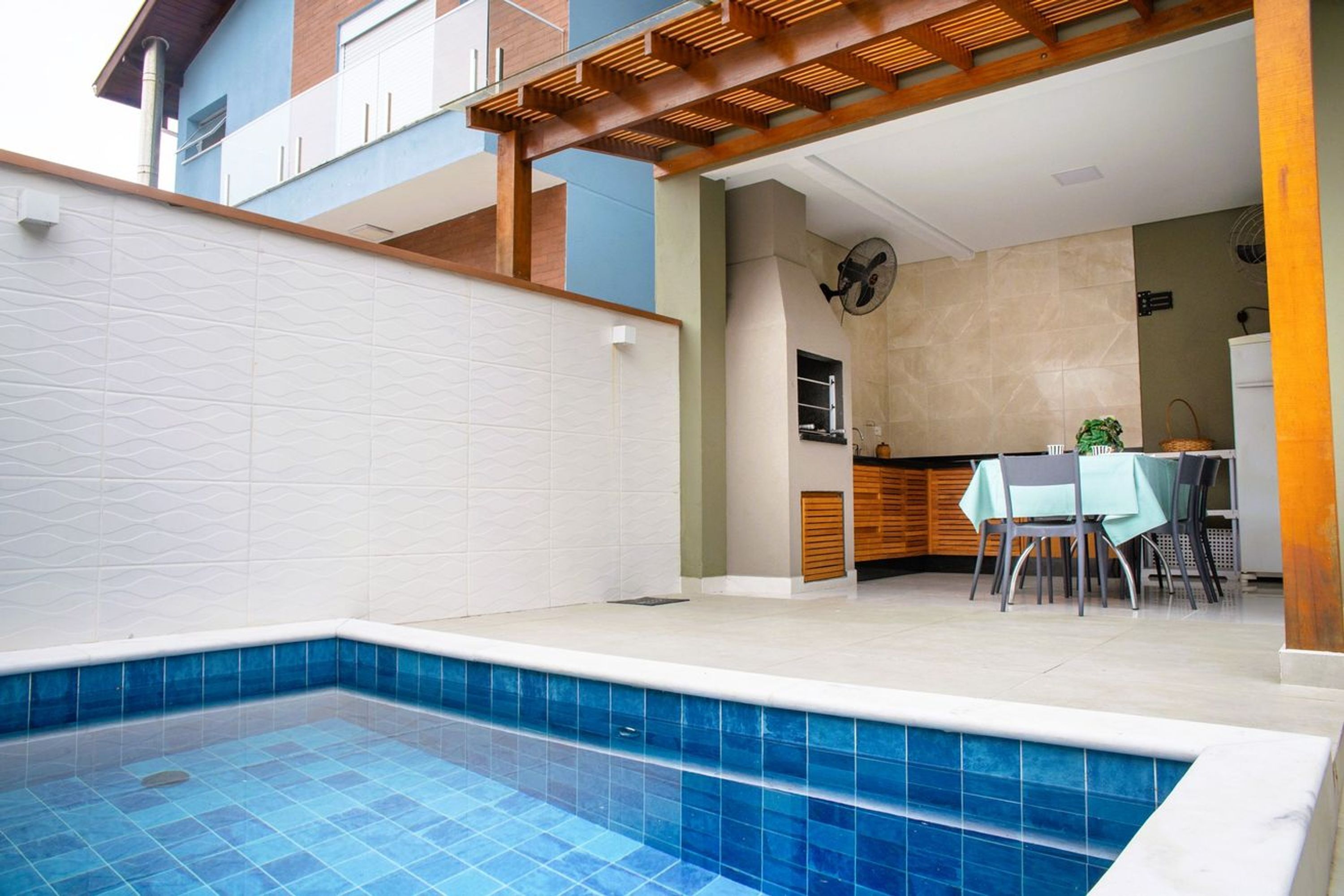 Piscina com borda azulejada, pergolado de madeira, área de refeições com mesas e cadeiras, e cozinha com geladeira.