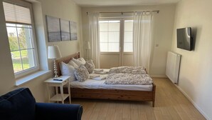 Quarto com cama de madeira, mesa de cabeceira com abajur, TV fixada na parede e janela com cortinas.