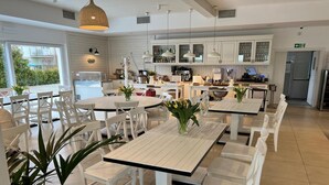 Uma área de refeições moderna com mesas e cadeiras brancas, uma cozinha ao fundo e vista para o exterior.