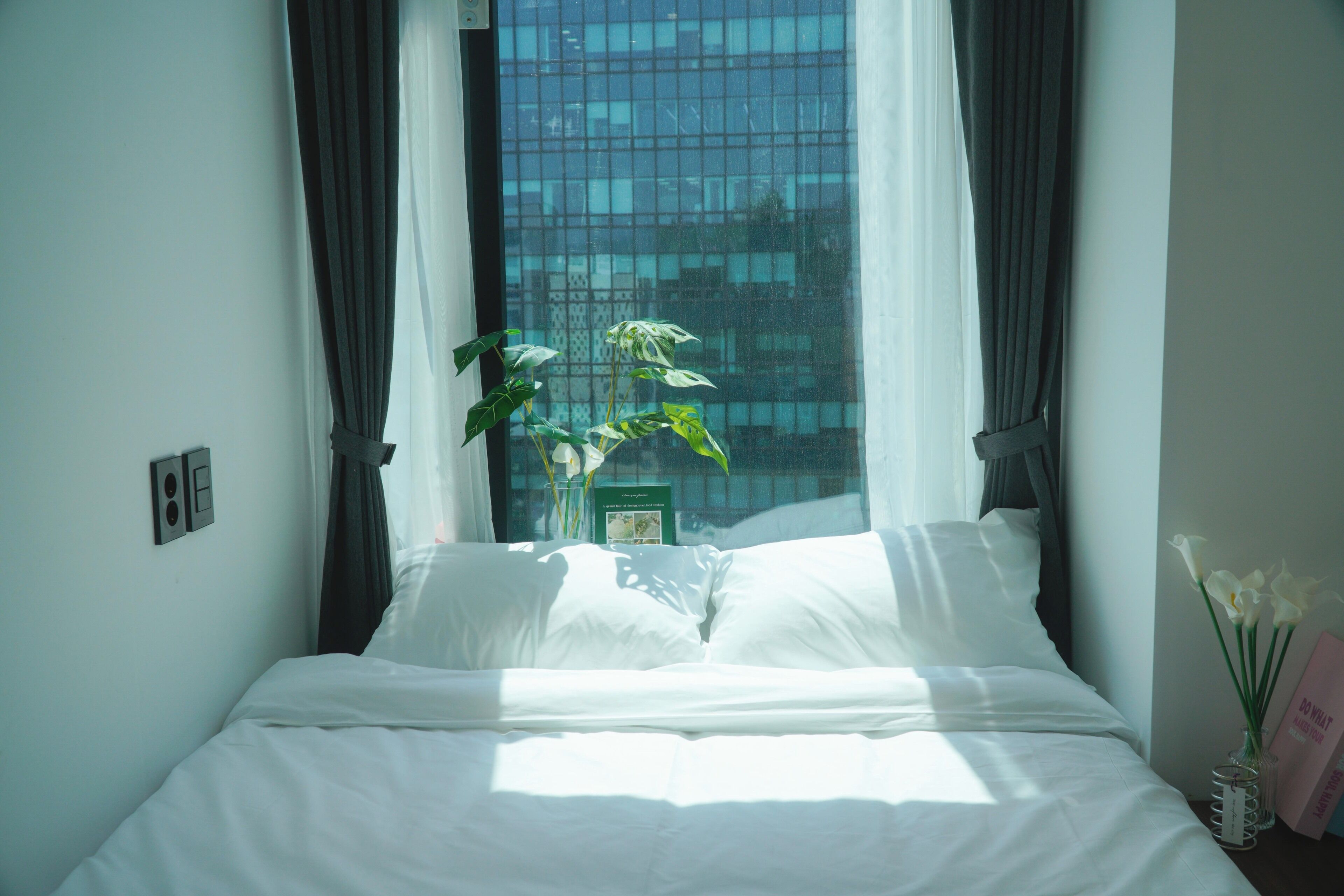 Quarto de hotel com uma cama, janela com cortinas, uma planta e um vaso com flores.