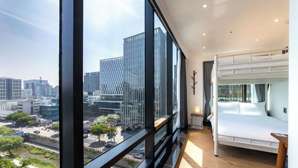 Quarto de hotel moderno com uma janela ampla, uma cama e vista para a paisagem urbana.