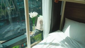 Uma cama com lençóis brancos, uma janela com vista para edifícios, um vaso com flores e um livro em uma mesa de cabeceira.