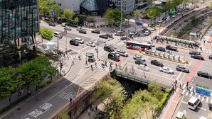 Paisagem urbana com edifícios altos e modernos, um cruzamento movimentado e uma faixa de pedestres.