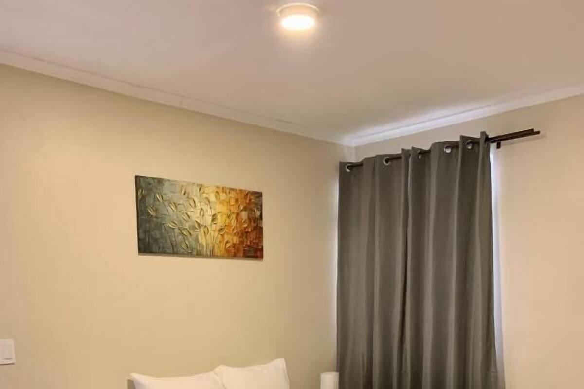 Quarto com cama, mesa de cabeceira, luminária, quadro na parede e cortina.