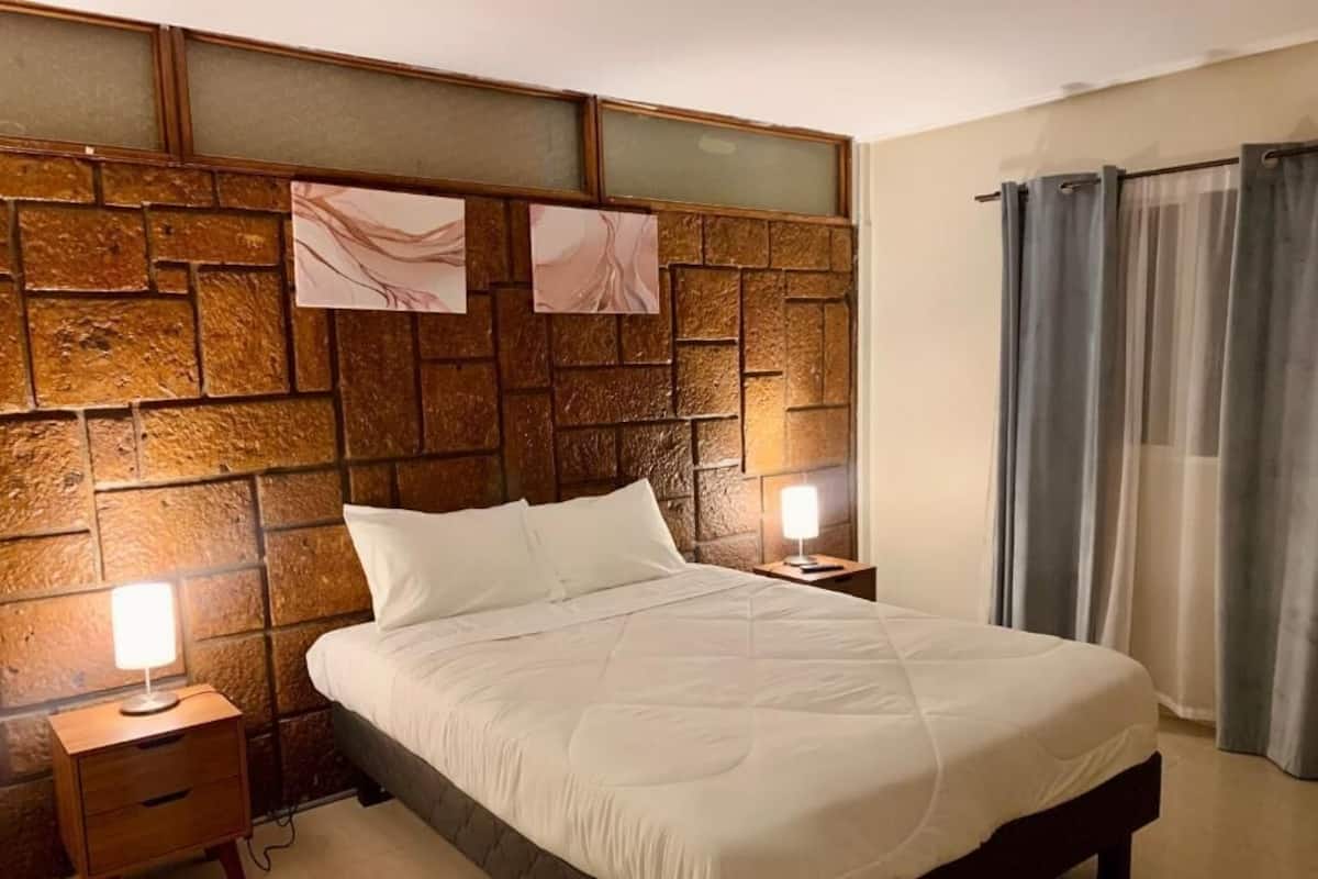 Quarto com cabeceira estampada em pedra, cama com roupa de cama branca, duas luminárias de cabeceira e uma janela com cortinas.