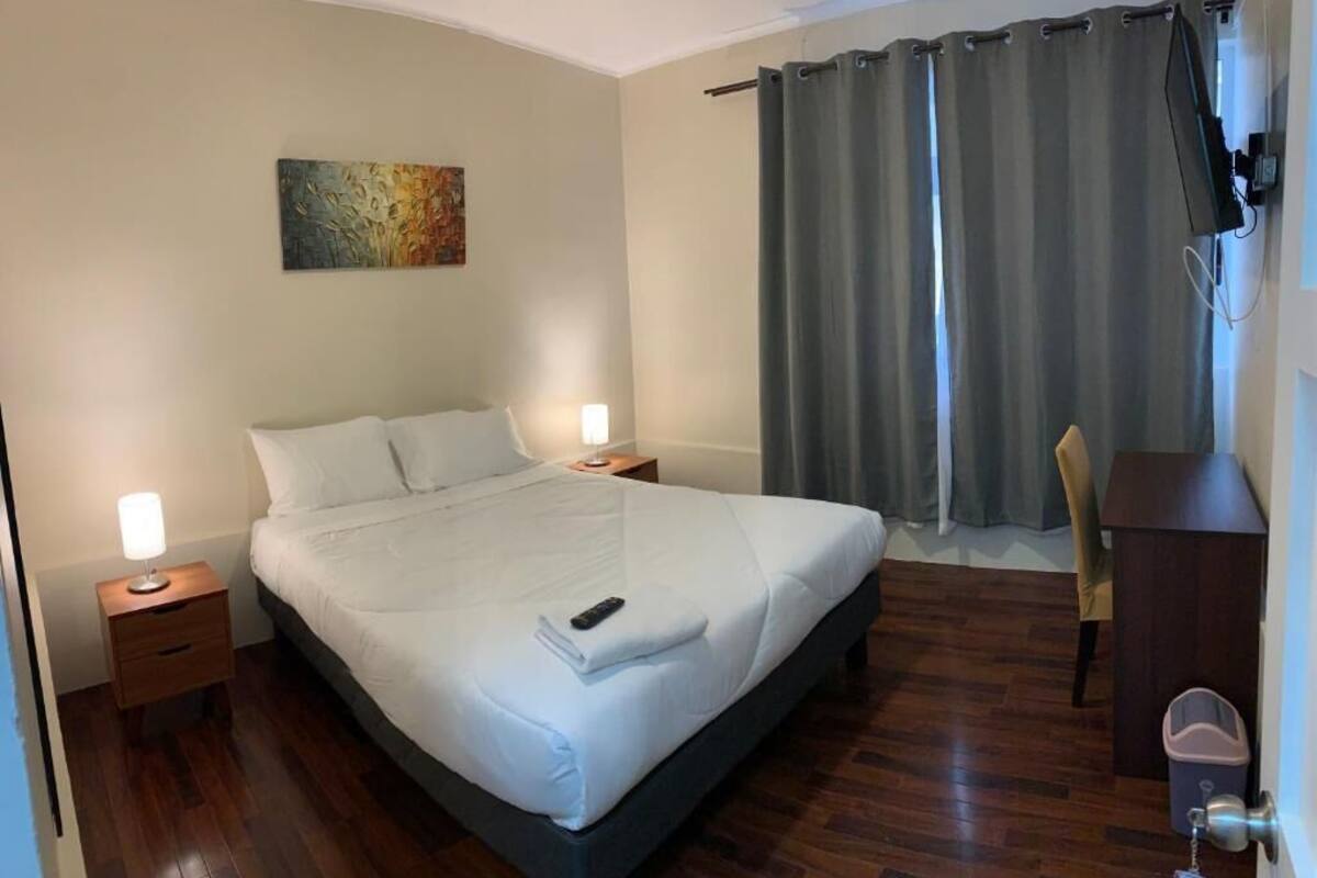 Quarto de hotel bem arrumado com cama, luminárias de cabeceira, escrivaninha e uma cadeira.