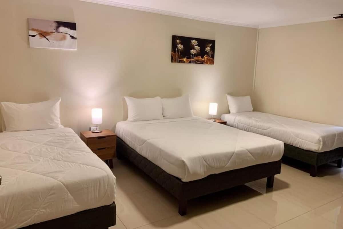 Quarto de hotel com três camas dispostas em linha, uma mesa de cabeceira com abajur e duas obras de arte penduradas na parede.