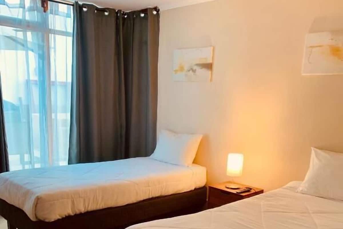 Quarto de hotel com duas camas, uma mesa de cabeceira com abajur, uma janela com cortinas e uma obra de arte na parede.