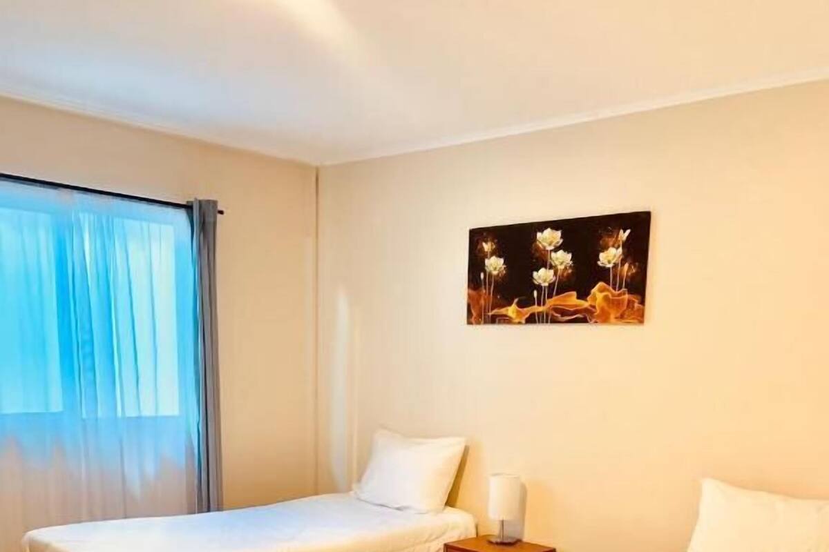 Quarto de hotel com duas camas de solteiro, uma cama grande, uma mesa de cabeceira com abajur e um quadro na parede.