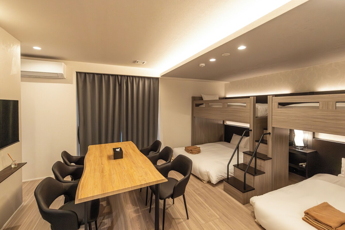 Quarto de hotel moderno com mesa de jantar de madeira, cadeiras pretas, beliche, escrivaninha com luminária e TV na parede.