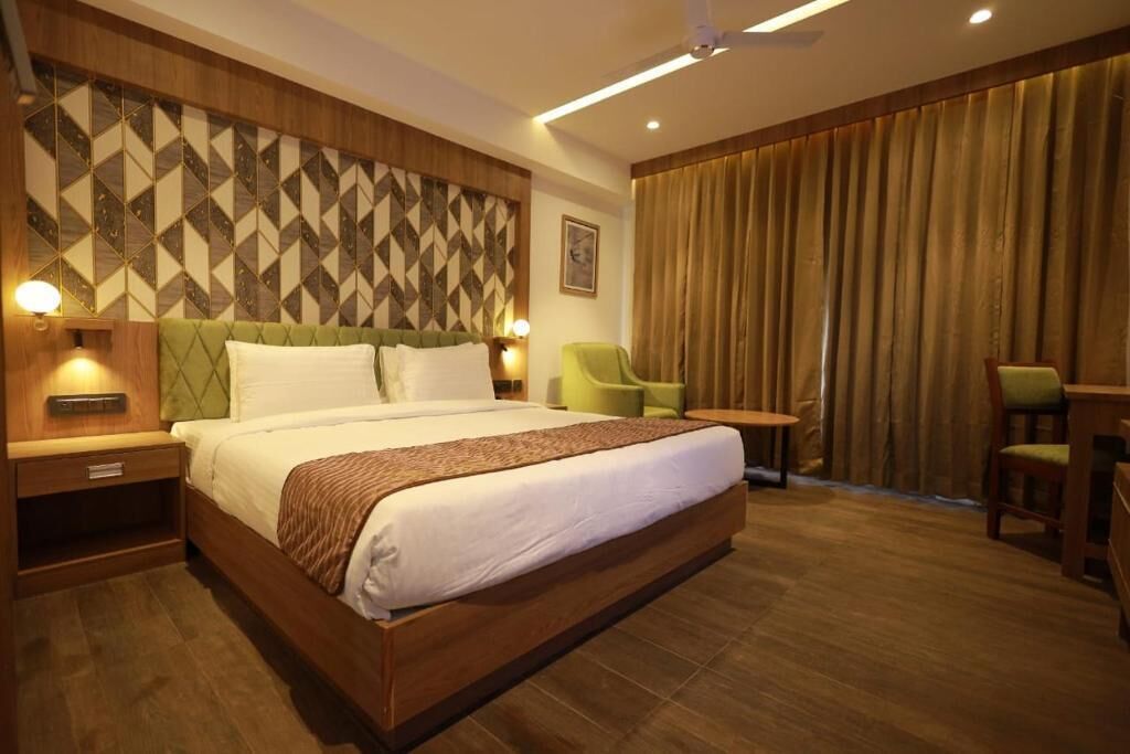 Quarto de hotel com uma cama grande, cabeceira com padrão geométrico, poltrona verde, uma mesinha e cortina.