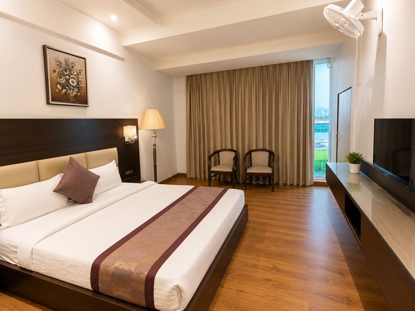 Quarto de hotel com cama, televisão, abajur, duas cadeiras e uma janela grande com cortinas.