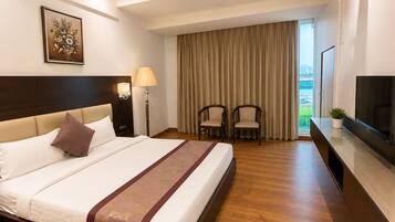 Quarto de hotel com cama, televisão, abajur, duas cadeiras e uma janela grande com cortinas.