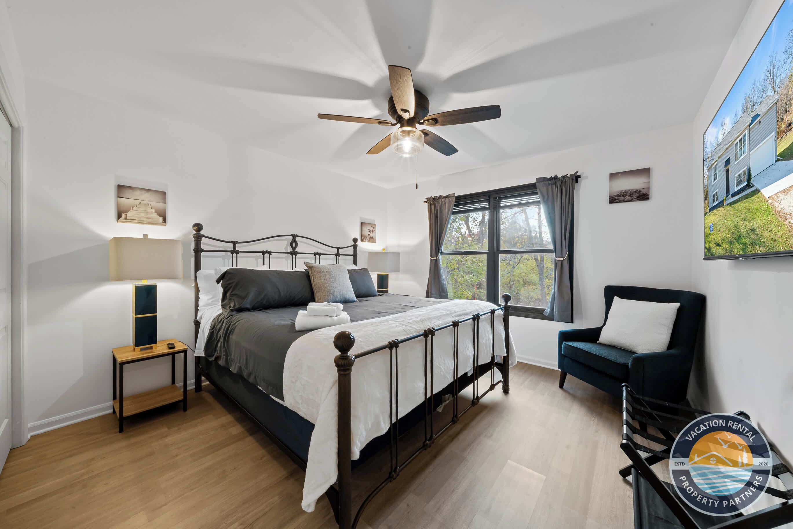 Quarto com cama, mesas de cabeceira, ventoinha no teto, janela com cortinas, cadeira e um quadro na parede.
