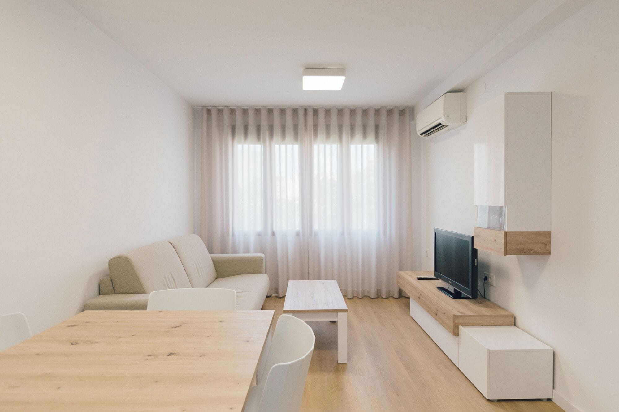 Uma sala de estar moderna e minimalista, com um sofá, uma mesa de centro de madeira, uma TV em um suporte de madeira e uma janela grande com cortinas leves.